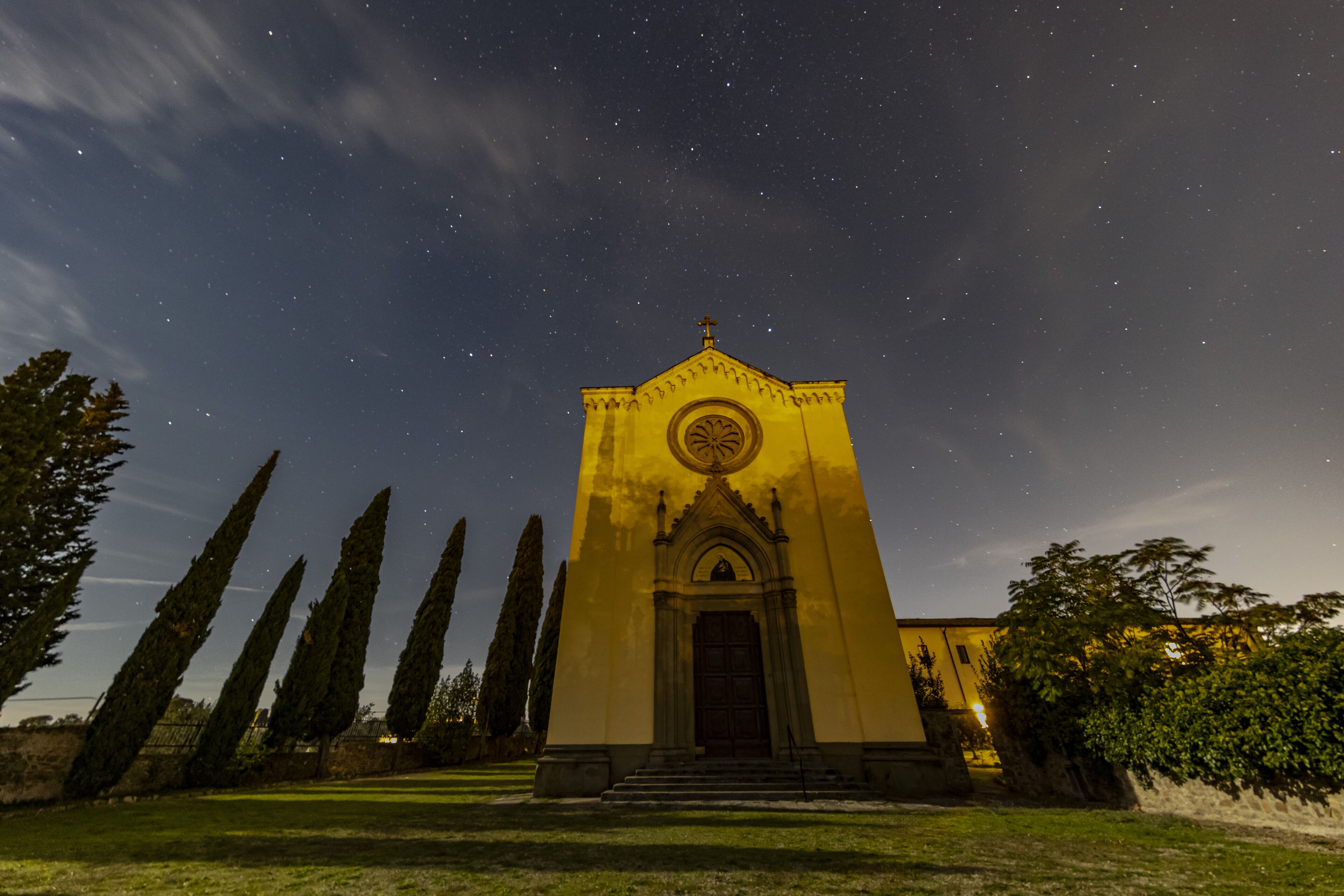 Uma igreja com uma janela circular notável e uma cruz no topo, cercada por um gramado bem cuidado e árvores altas.