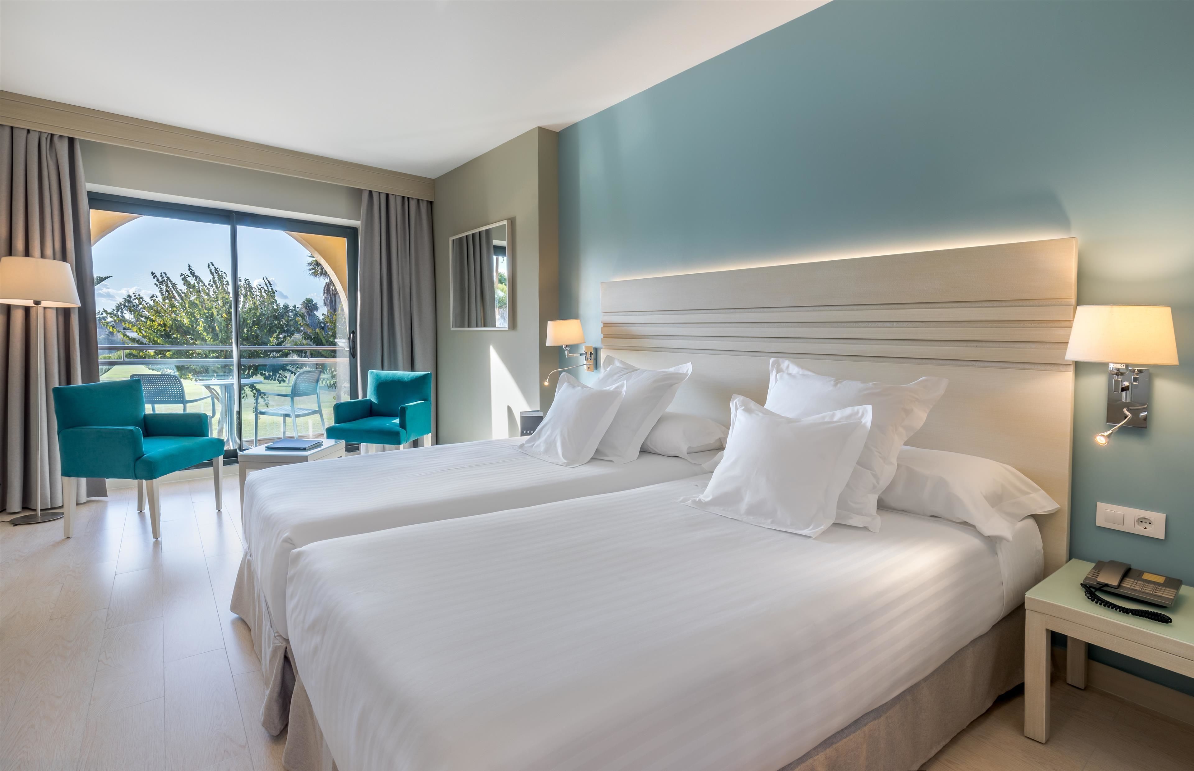 Quarto de hotel com duas camas, uma varanda com cadeiras e vista para o verde.
