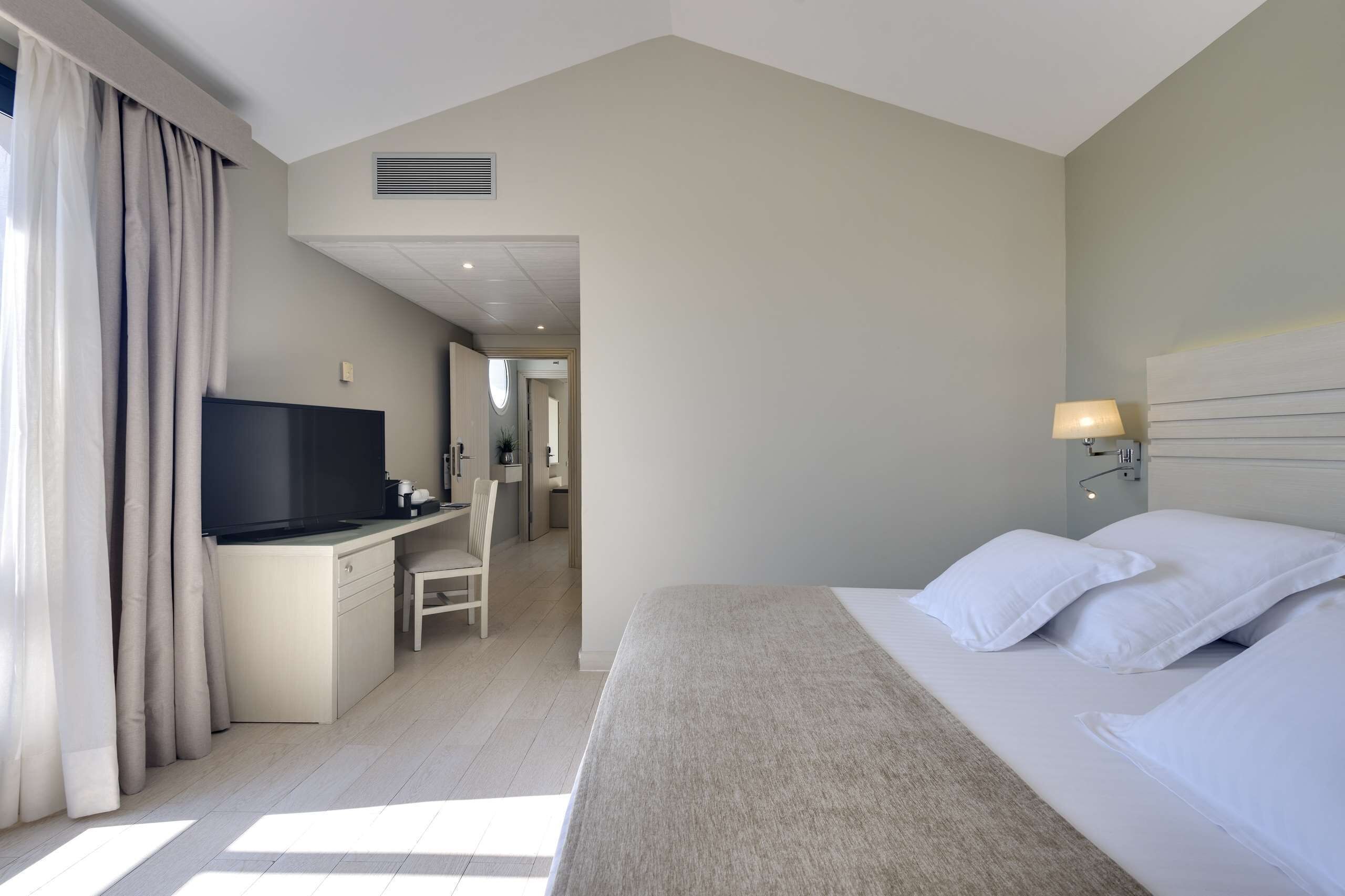 Quarto de hotel com cama, secretária, televisão e abajur.