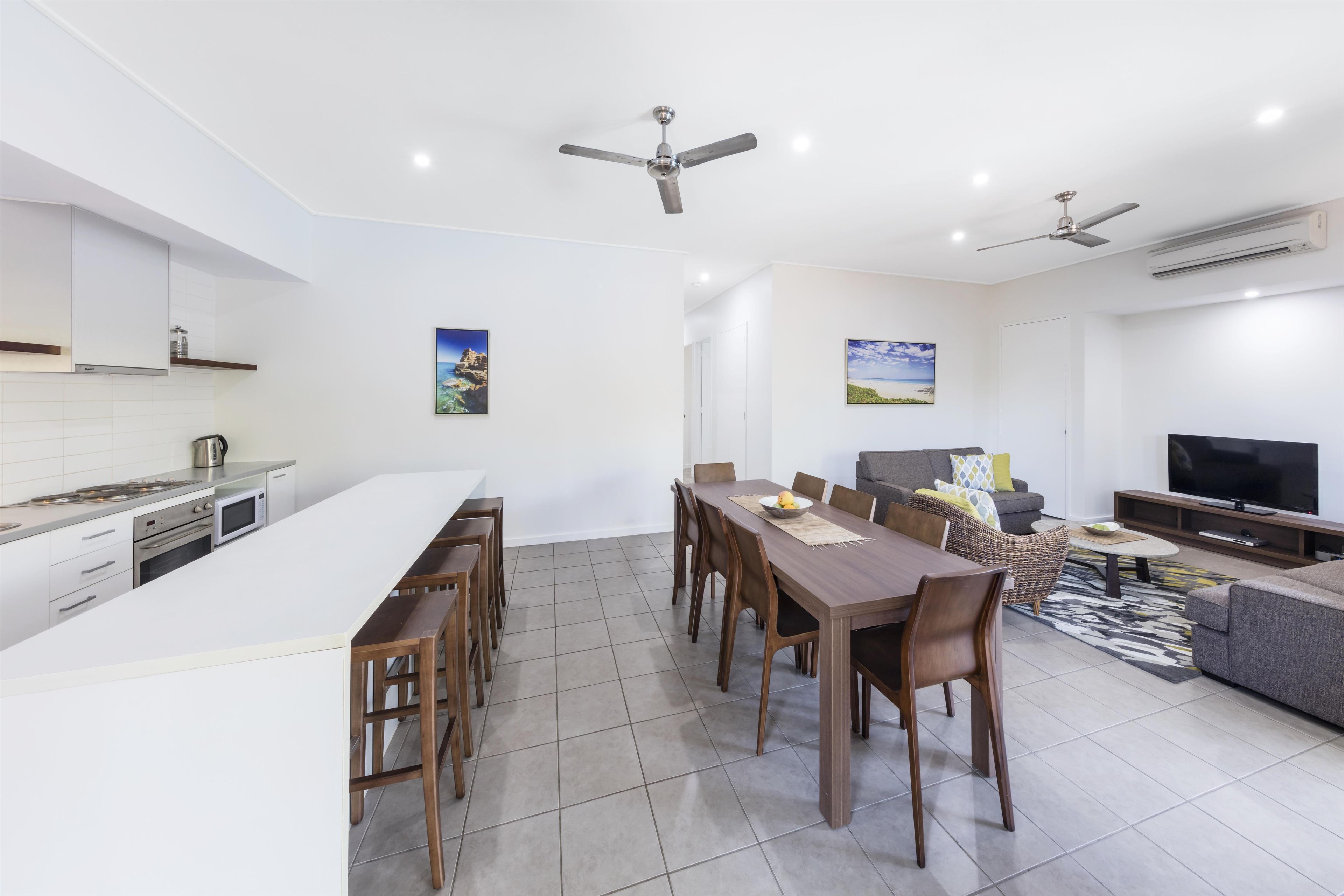 Cozinha moderna com ilha central, área de jantar com cadeiras de madeira, sala de estar com sofá e TV, e ventilador de teto.