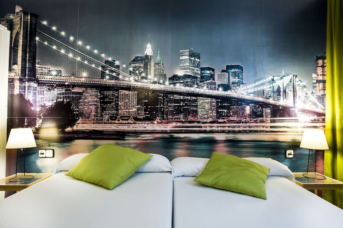 Quarto de hotel com uma cama, duas almofadas, duas mesas de cabeceira com luminárias e um grande mural da paisagem urbana na parede.