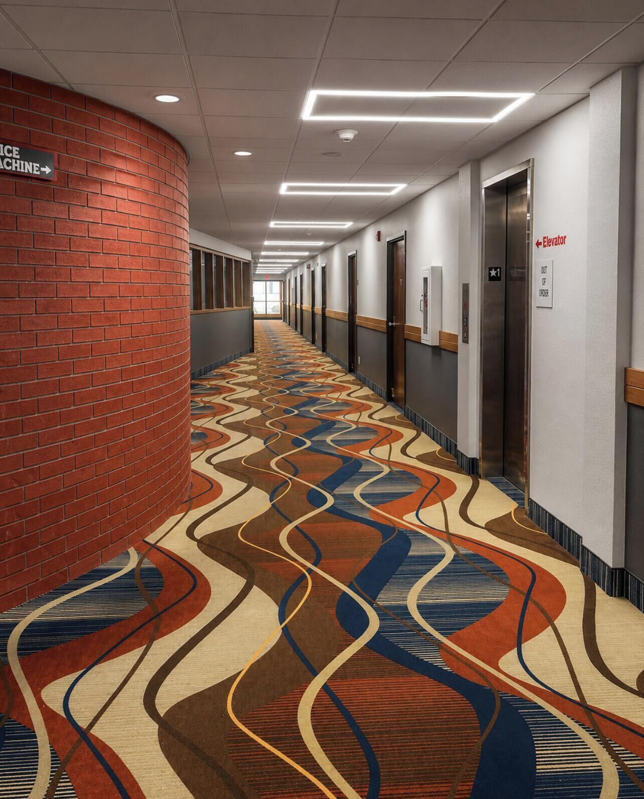 Corredor de hotel com carpete estampado, parede de tijolos e elevador.