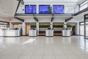Um hall amplo com design moderno, destacando um balcão de recepção central, um balcão no segundo andar com cortinas azuis e uma parede verde com plantas.