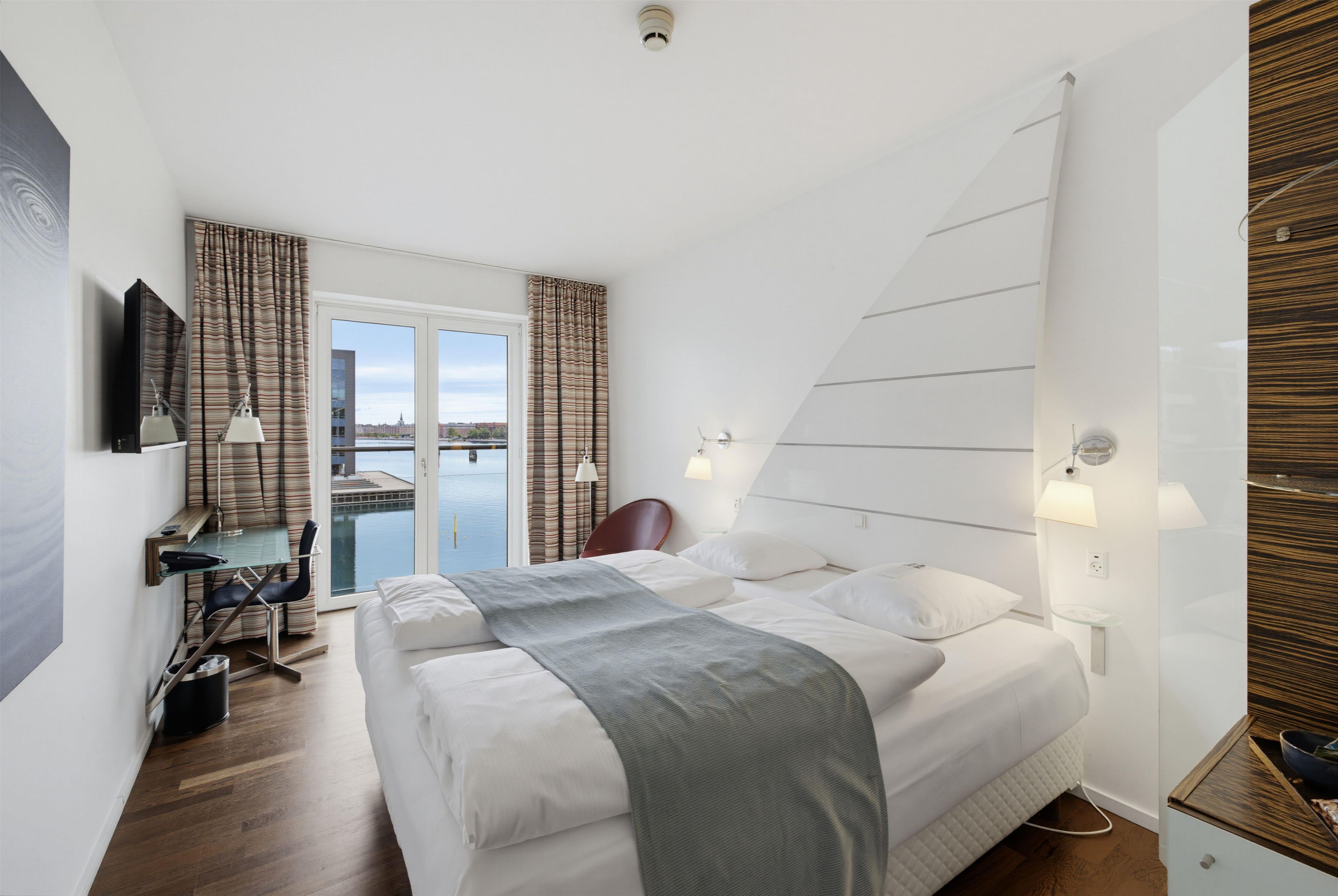 Quarto de hotel com cama, escrivaninha, cadeira e vista da cidade pela janela.