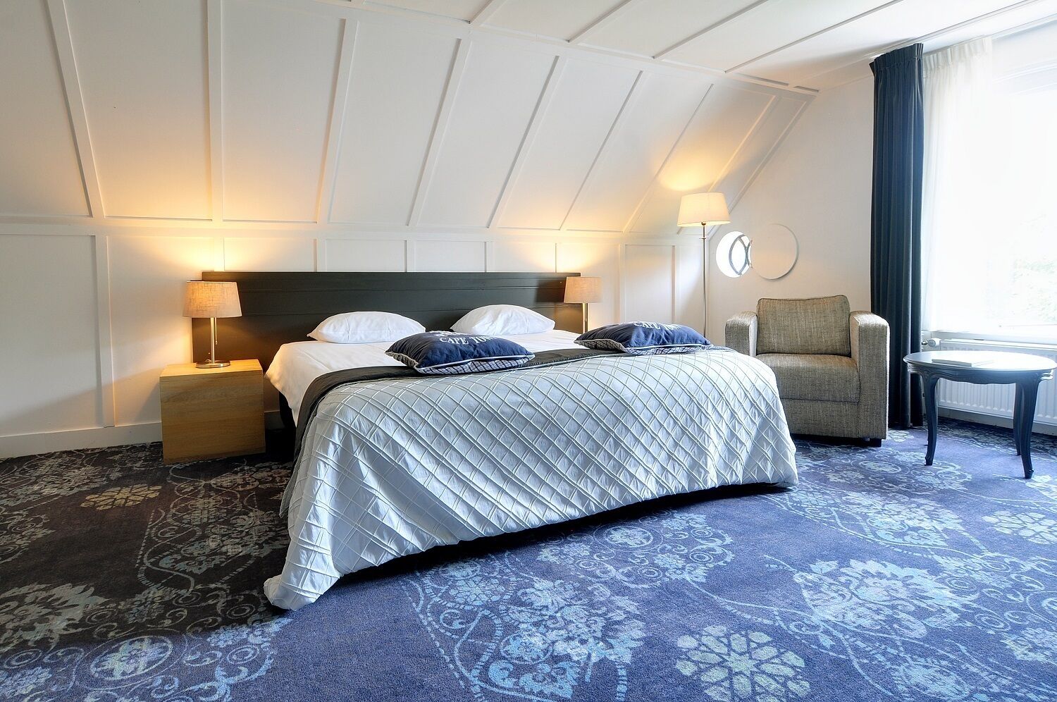 Quarto de hotel com uma cama grande, duas mesas de cabeceira com abajures, uma cadeira e uma pequena mesa.