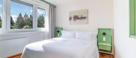 Quarto de hotel com uma cama grande, janela com cortinas, um quadro na parede e uma mesa de cabeceira verde.