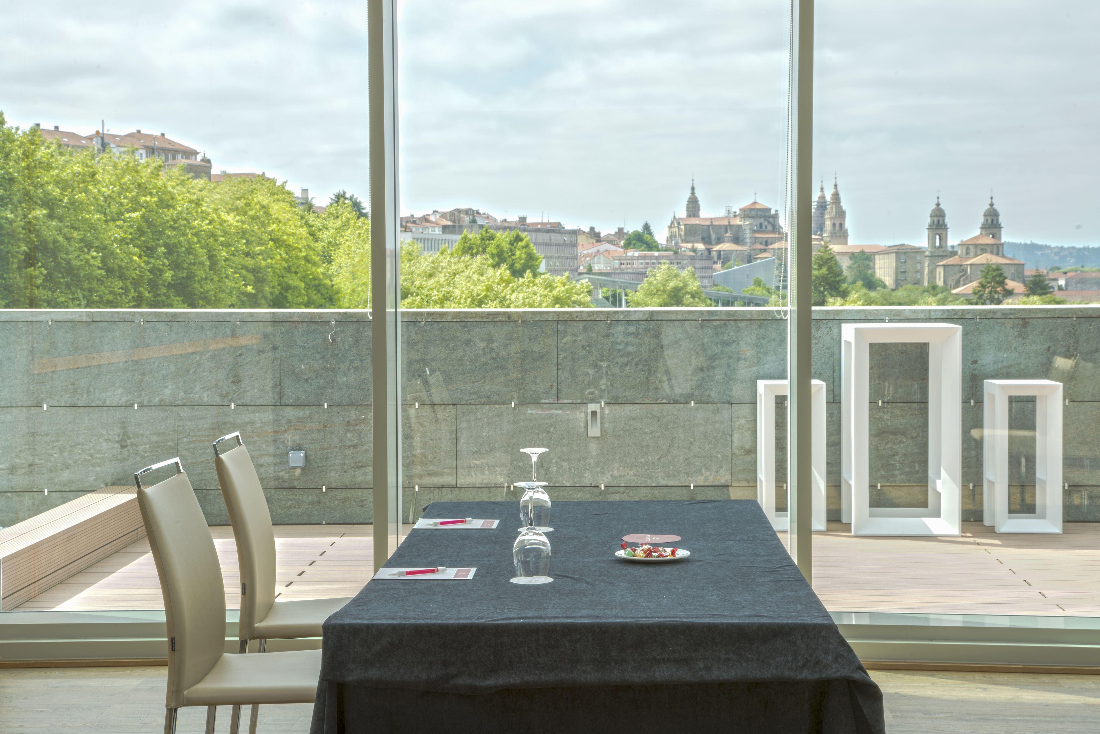 Uma área de refeições com uma mesa posta para dois, um copo de água e um prato de comida. A mesa tem como plano de fundo um horizonte urbano com edifícios históricos e árvores.