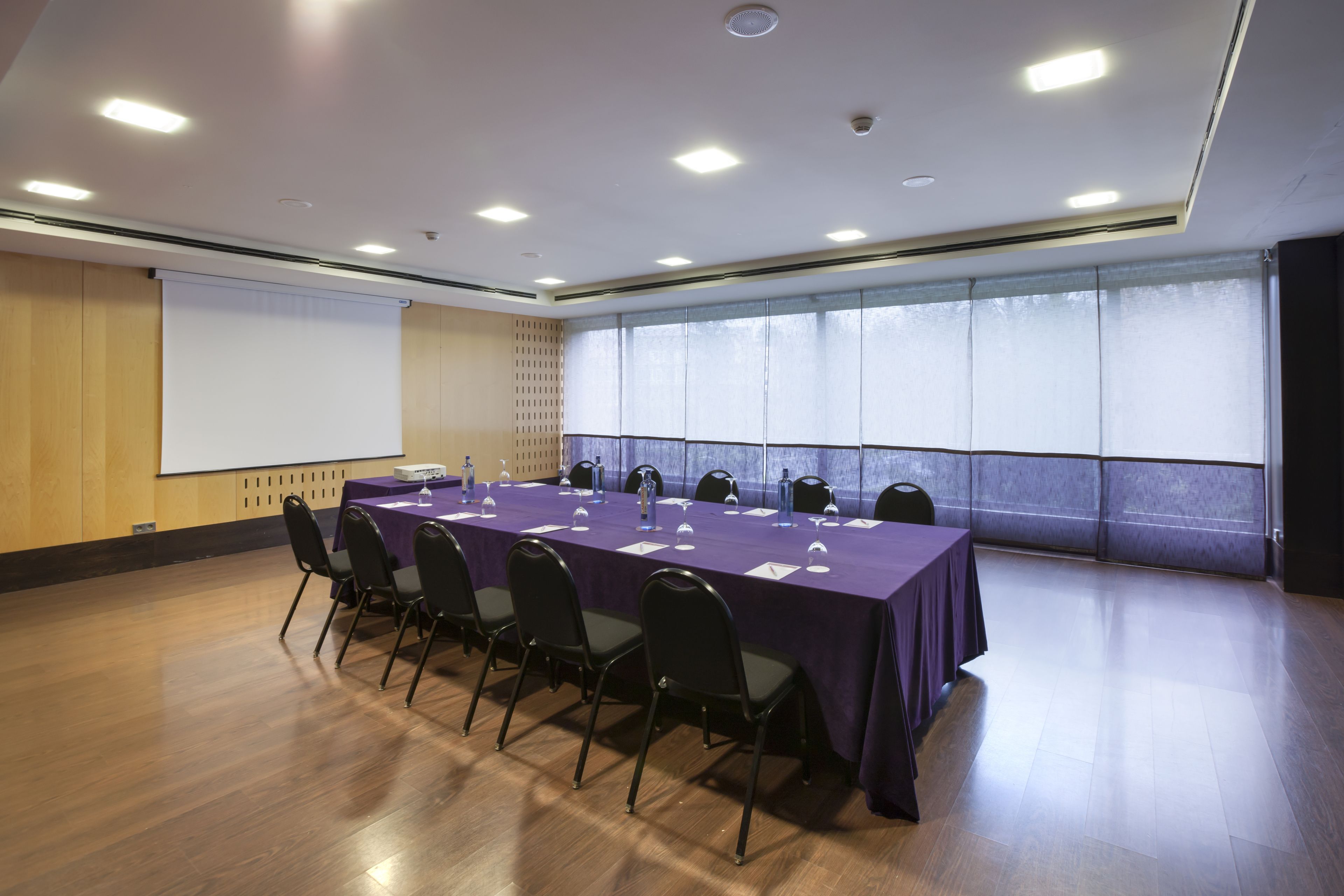 Uma sala de conferências com uma mesa comprida, cadeiras, tela de projetor e uma janela grande com persianas.