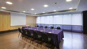 Uma sala de conferências com uma mesa comprida, cadeiras, tela de projetor e uma janela grande com persianas.