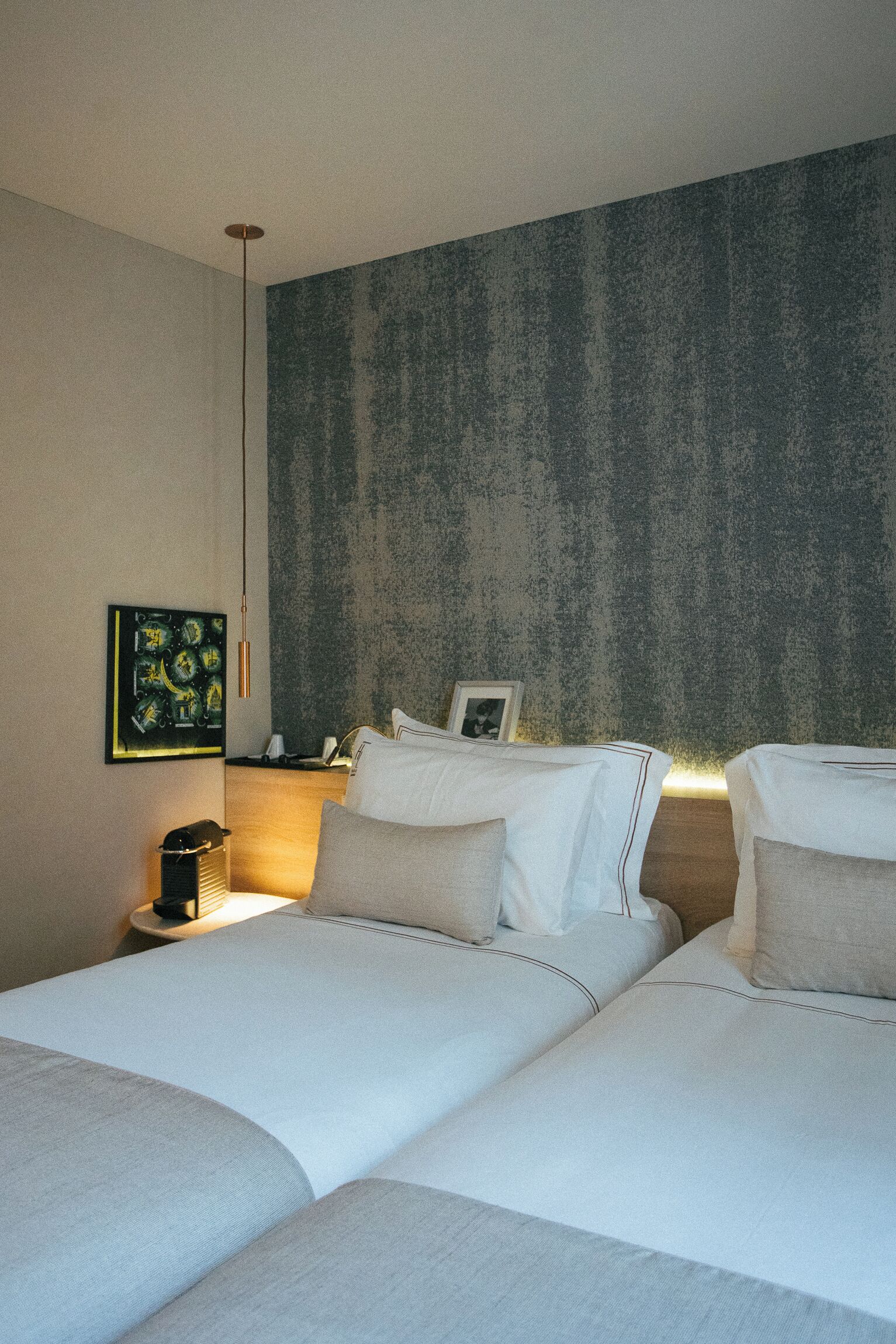 Quarto de hotel com duas camas, uma mesinha de cabeceira com abajur, um quadro na parede e parede texturizada atrás das camas.