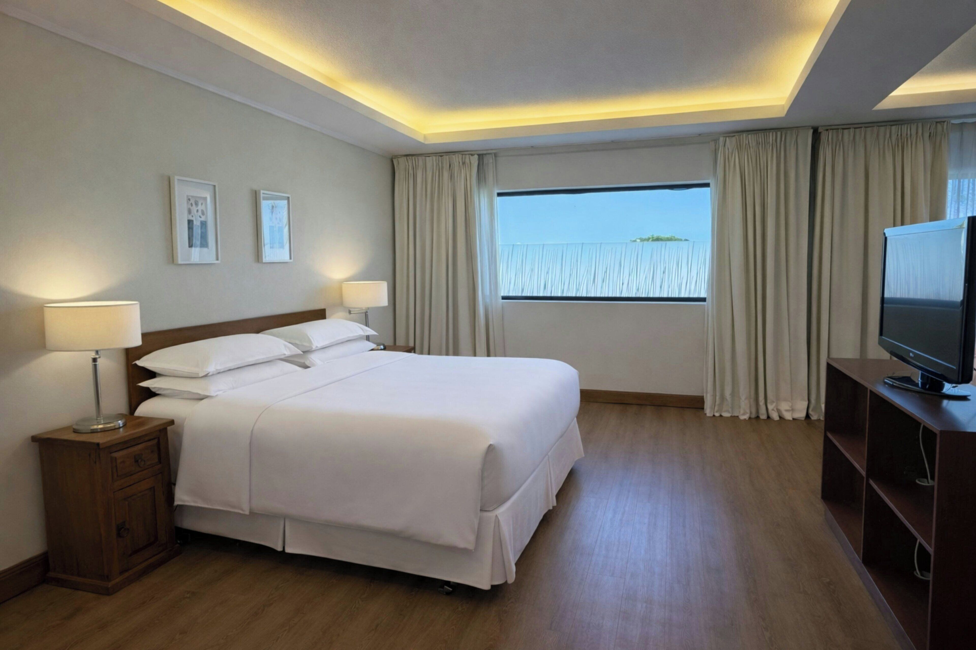 Quarto de hotel com uma cama grande, duas luminárias de cabeceira, uma TV sobre um suporte e uma janela com cortinas.