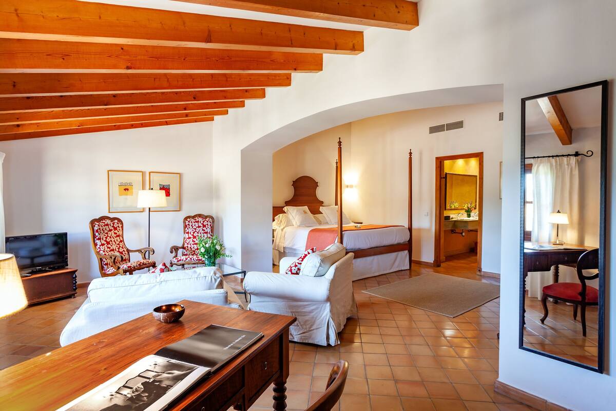 Quarto de hotel com teto de madeira, uma cama, uma escrivaninha com cadeira, televisão e espelho.