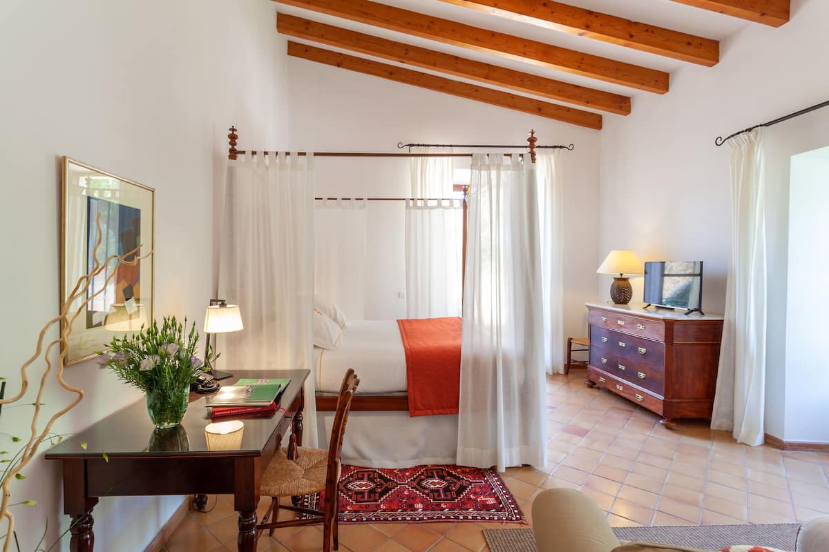 Quarto com cama, mesa de cabeceira com abajur, cômoda, cadeira e janela com cortinas.