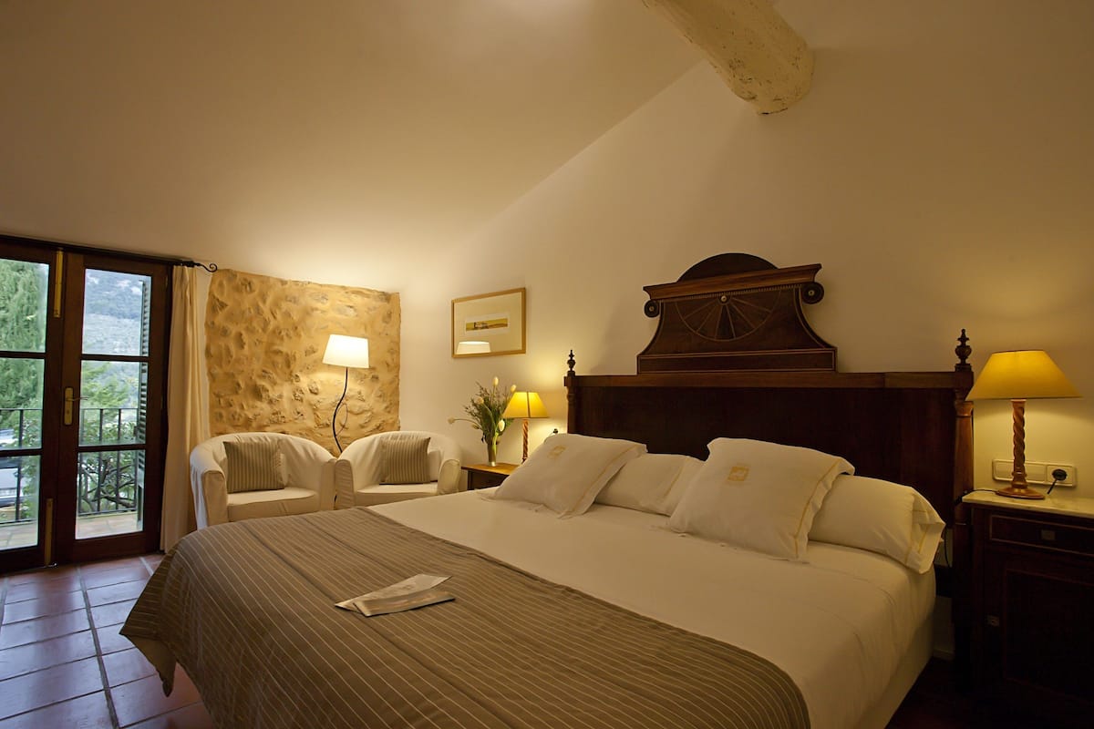 Quarto de hotel com uma cama grande, duas mesas de cabeceira com abajures, uma cadeira, uma pequena mesa com um vaso e uma janela com cortinas.