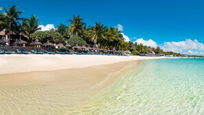 Uma praia com águas turquesa cristalinas, areia branca e palmeiras. Há espreguiçadeiras e guarda-sóis ao longo da costa.