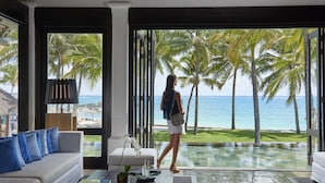 Sala de estar moderna com uma grande porta de vidro que dá para uma área de piscina, com vista para uma praia com palmeiras e o oceano.