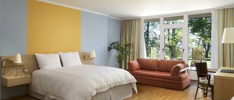 Quarto de hotel com uma cama grande, um sofá vermelho, uma escrivaninha com cadeira e uma janela com cortinas.