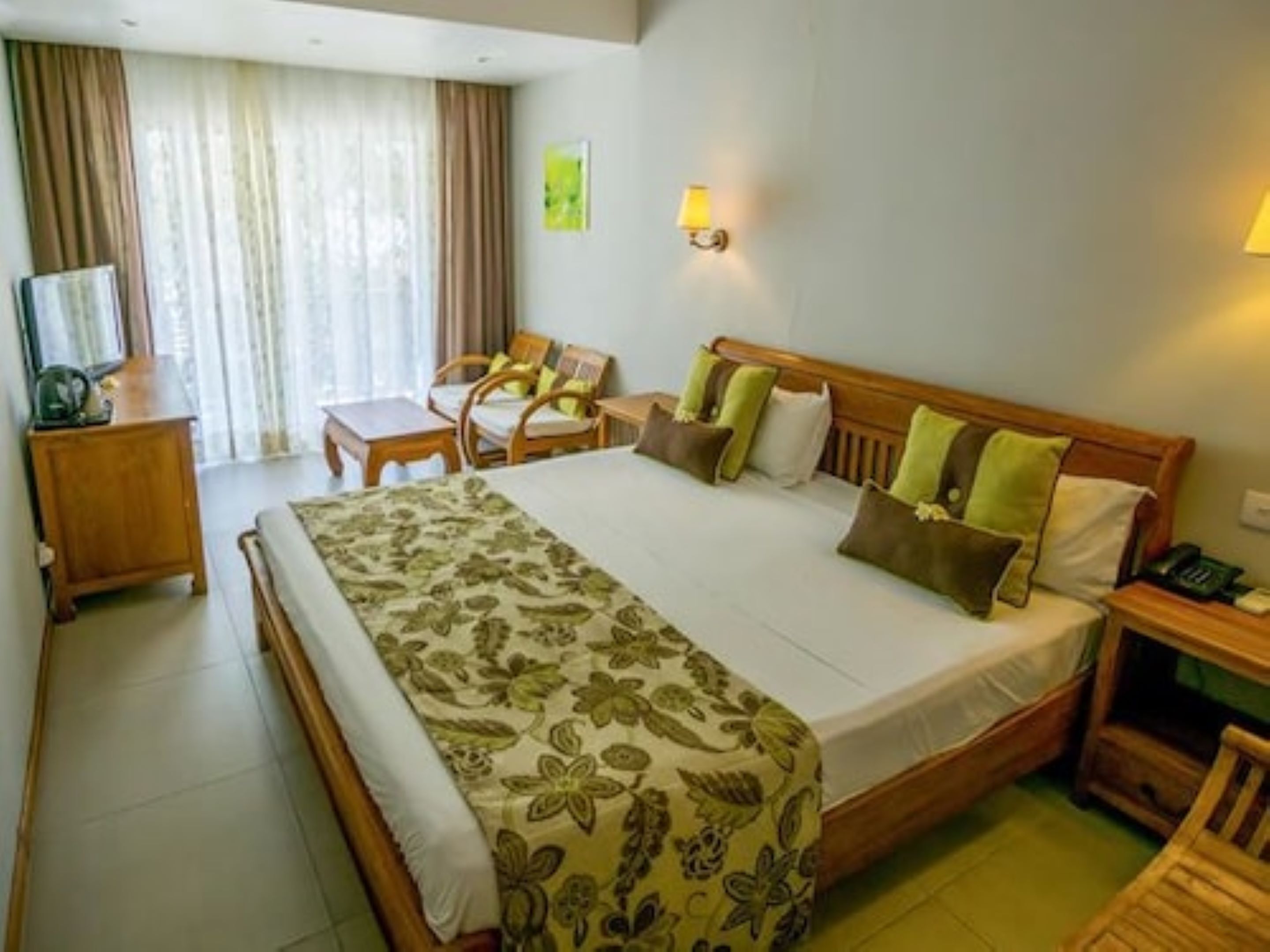 Quarto de hotel com uma cama grande, cabeceira de madeira, uma escrivaninha com cadeira, uma mesinha e uma televisão sobre um móvel de madeira.