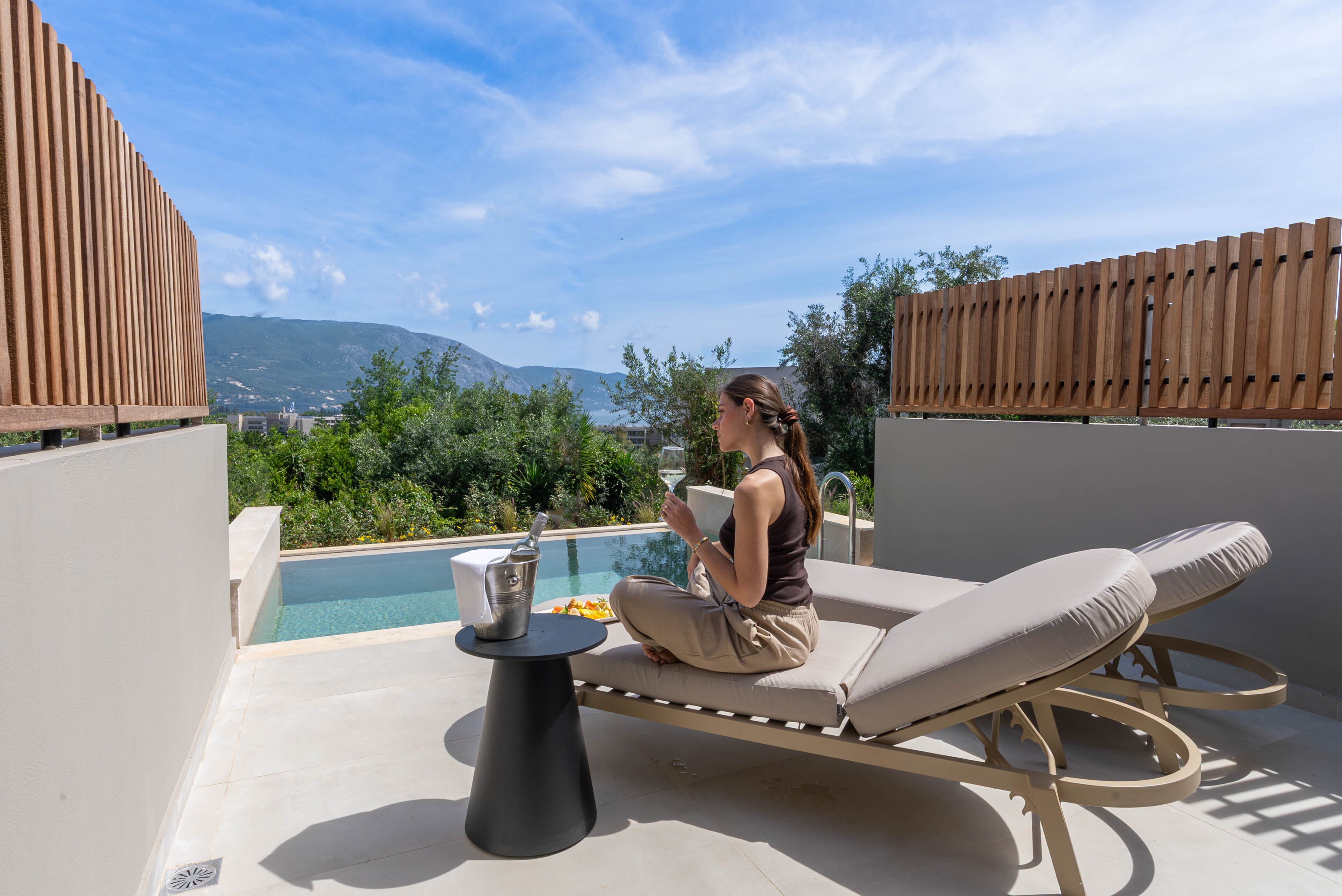 Pessoa sentada em espreguiçadeira perto de uma piscina, com vista para montanhas e vegetação.