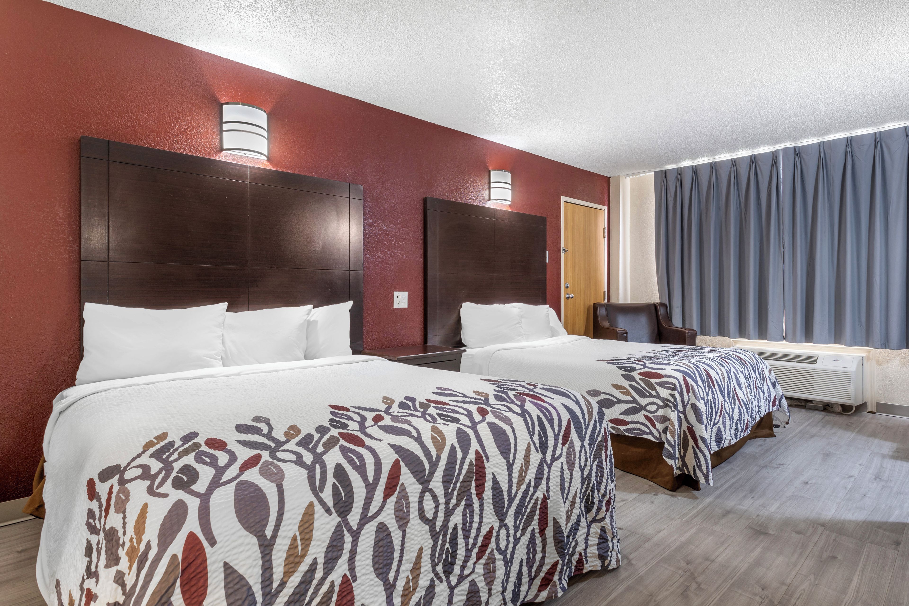 Quarto de hotel com duas camas, cabeceira de madeira, janela com cortinas e luminária fixada na parede.
