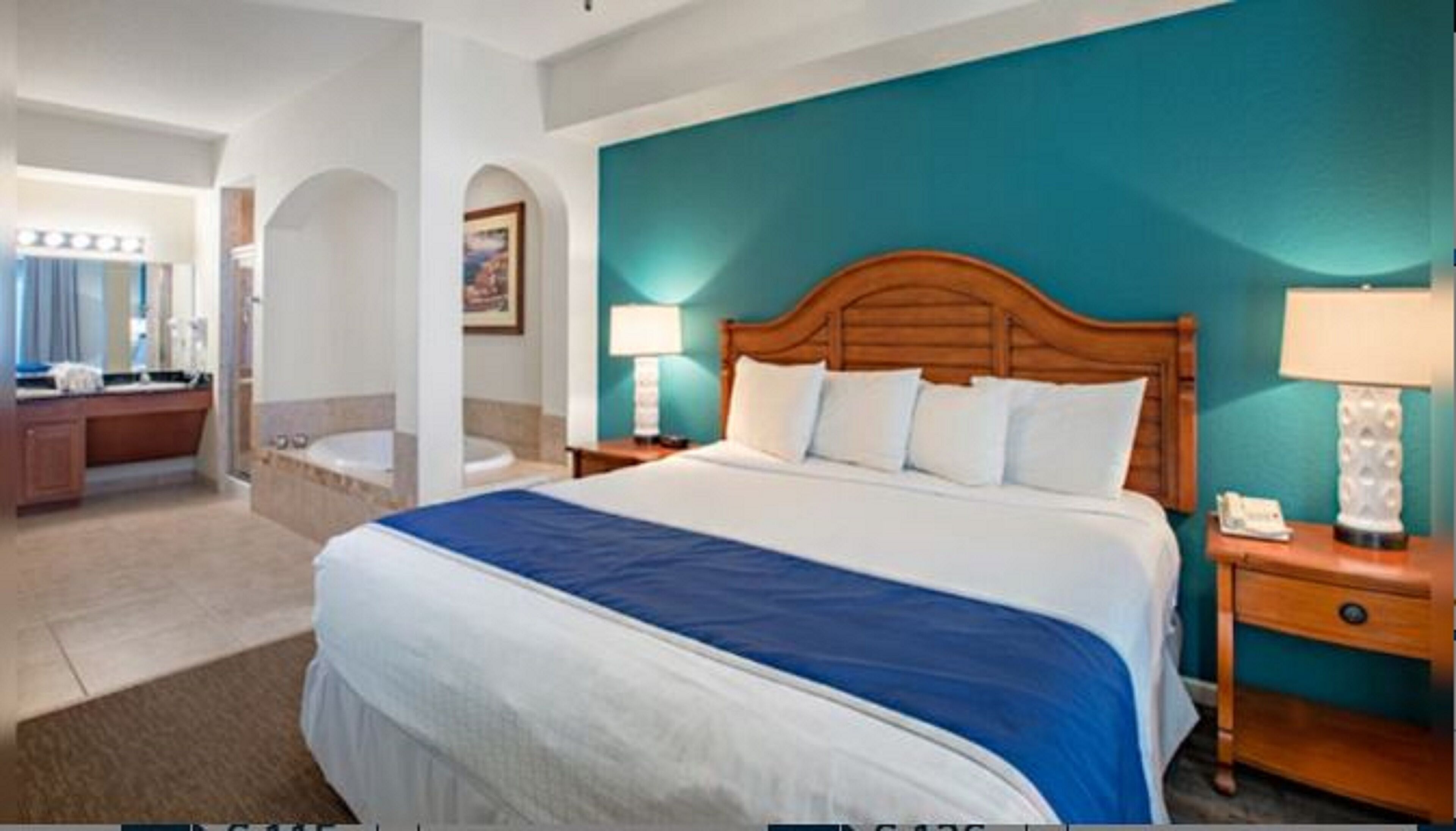 Quarto de hotel com uma cama grande, cabeceira de madeira, duas mesas de cabeceira com abajures, banheiro com banheira e uma escrivaninha com cadeira.