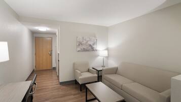 Uma sala de estar moderna com um sofá, uma mesa de centro, uma poltrona e um abajur. Há uma porta que dá para outro cômodo e um quadro na parede.
