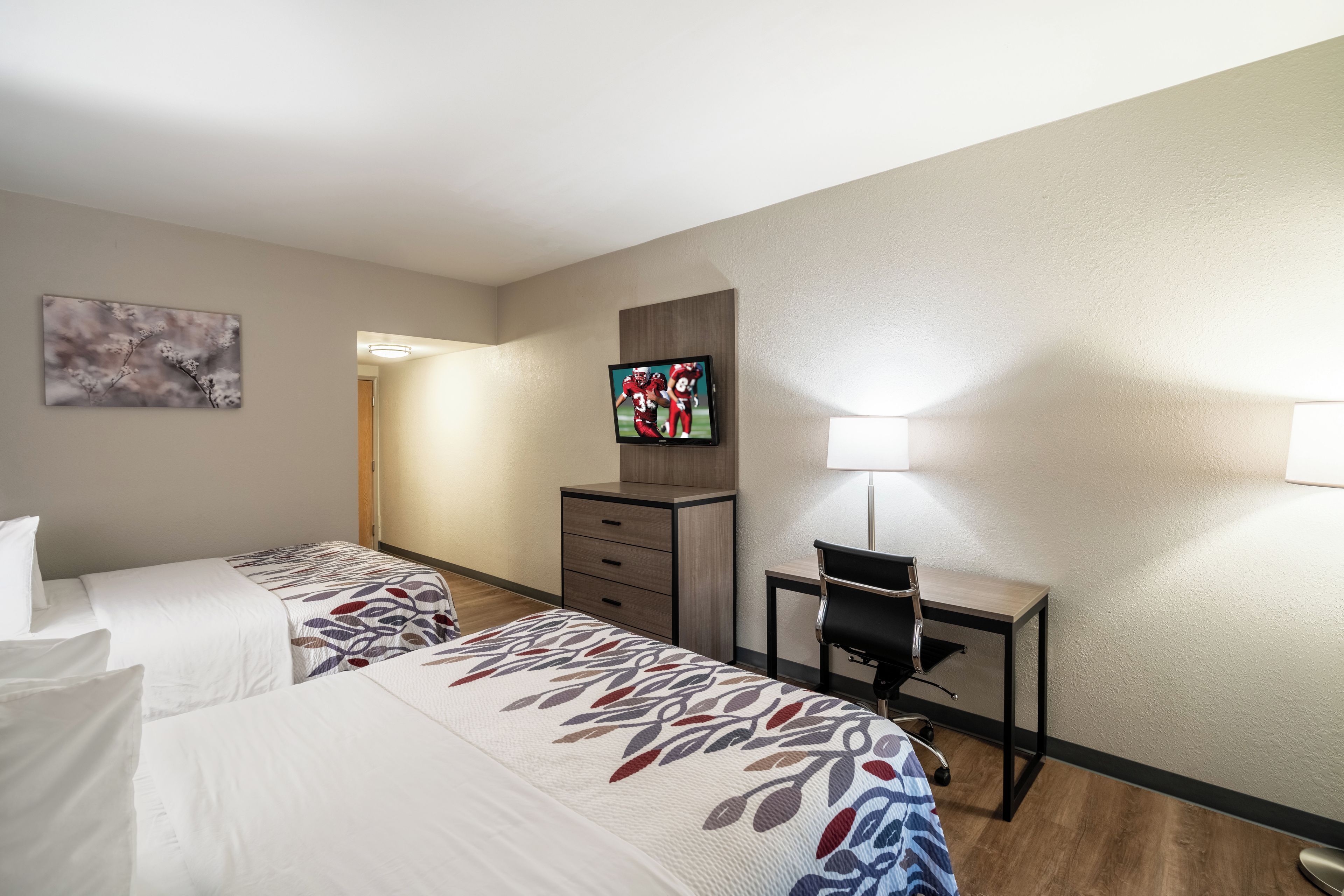 Quarto de hotel com duas camas de solteiro, uma mesa de trabalho com cadeira, televisão e luminária.