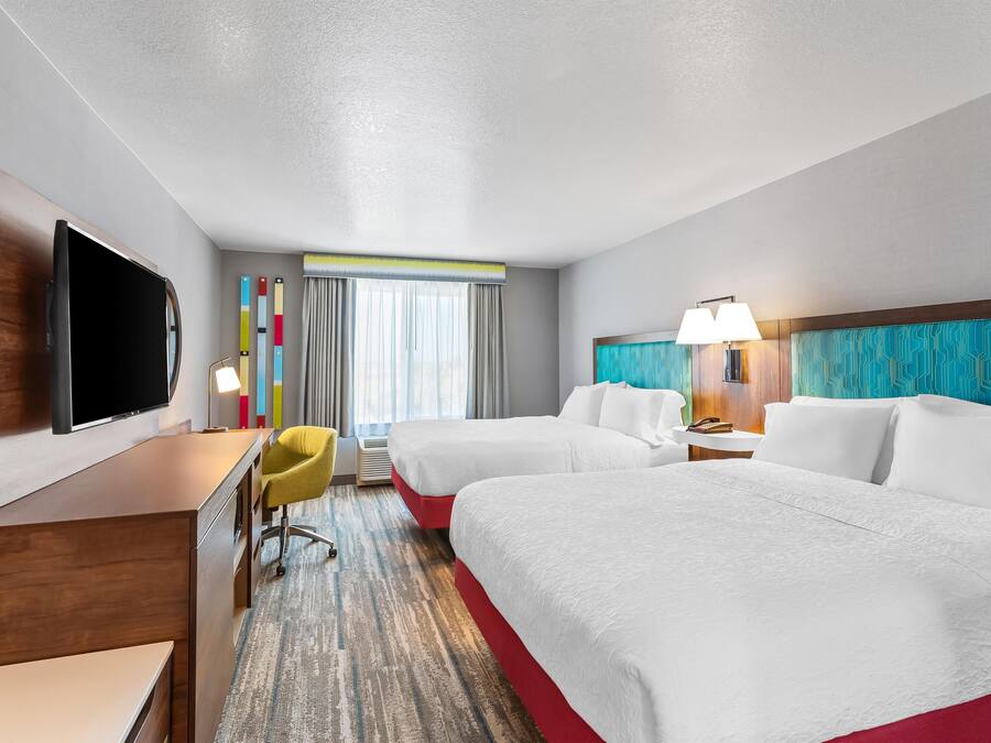 Quarto de hotel com duas camas, uma escrivaninha, uma televisão, uma cadeira e uma janela com cortinas.