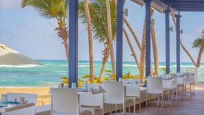 Um restaurante à beira-mar com cadeiras e mesas brancas, palmeiras e vista para o oceano.