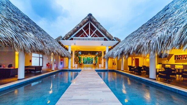Um resort com piscina central, estruturas com telhados de palha e uma galeria coberta.