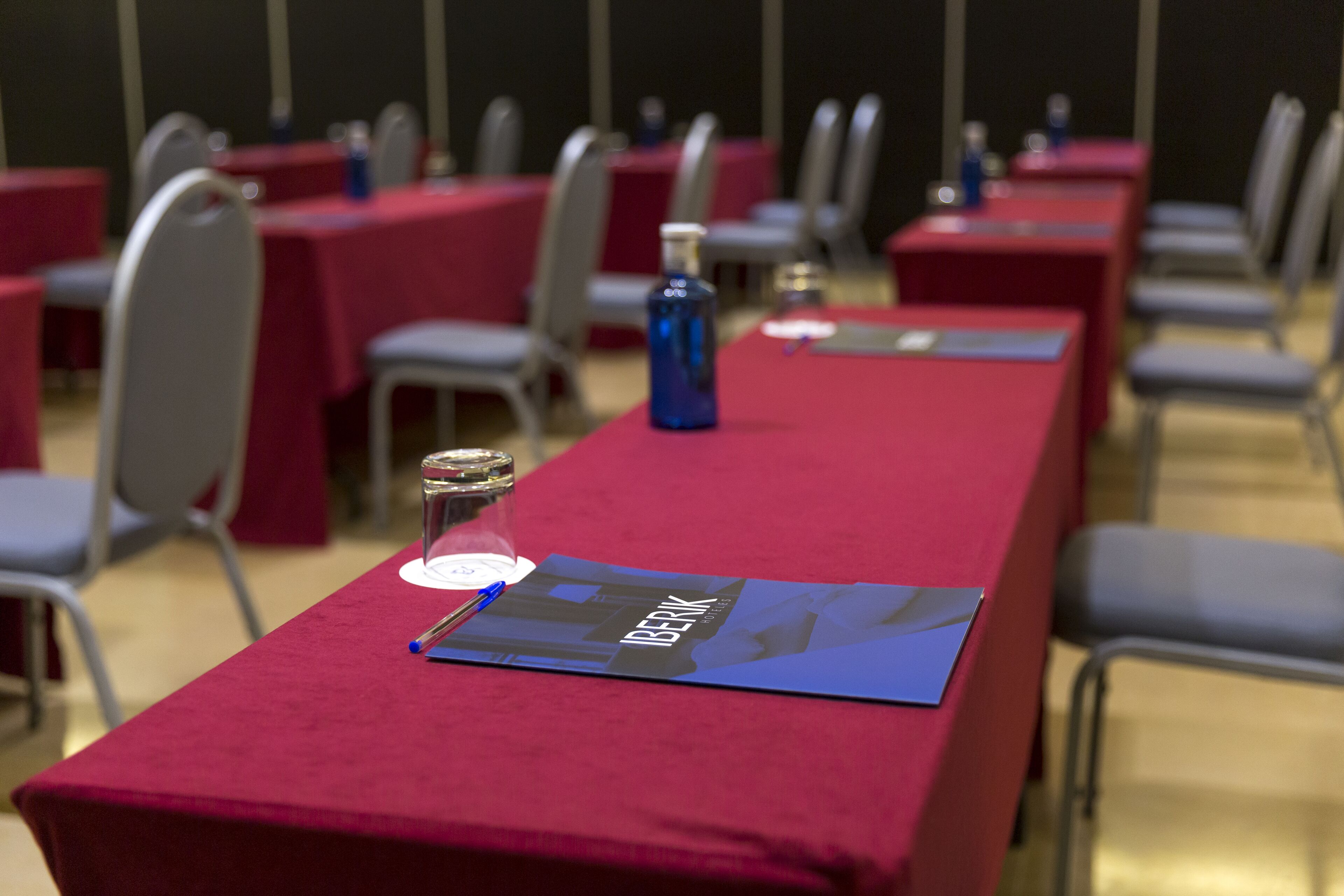 Uma sala de conferências com mesas cobertas por toalhas vermelhas, cadeiras e uma garrafa azul sobre uma das mesas.