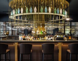 Área de bar com lustre central, diversas garrafas de bebidas alcoólicas e banquetas.