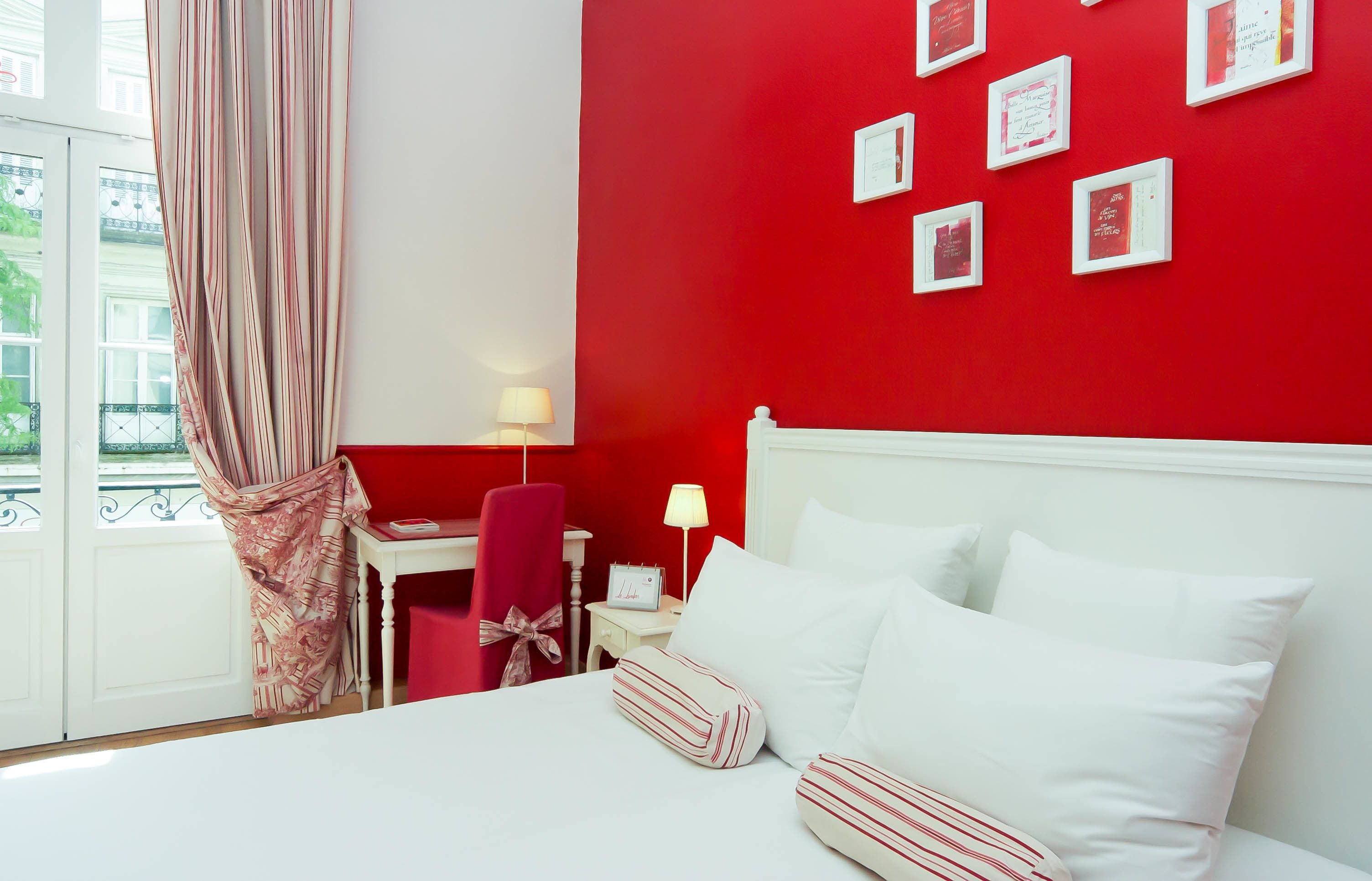 Quarto de hotel com uma parede de destaque vermelha, uma cama branca e uma mesa com luminária.