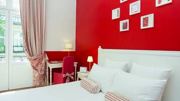 Quarto de hotel com uma parede de destaque vermelha, uma cama branca e uma mesa com luminária.