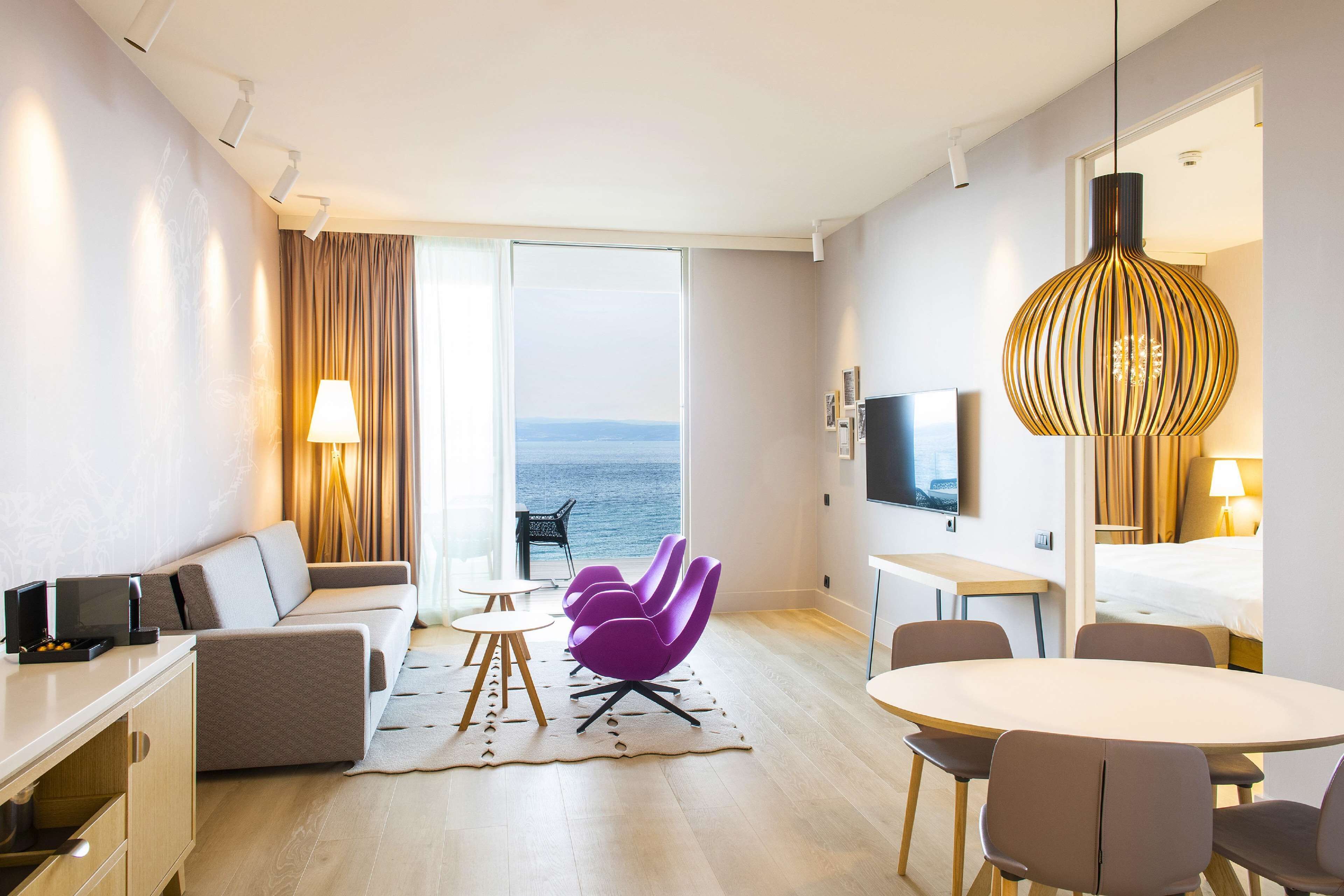 Quarto de hotel moderno com sofá, cadeiras roxas, mesa de jantar e televisão.