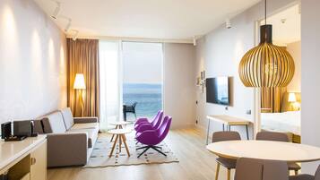 Quarto de hotel moderno com sofá, cadeiras roxas, mesa de jantar e televisão.