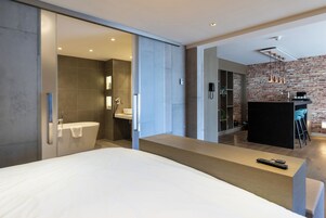 Quarto de hotel moderno com uma cama grande, banheiro com banheira e área de cozinha com balcão.