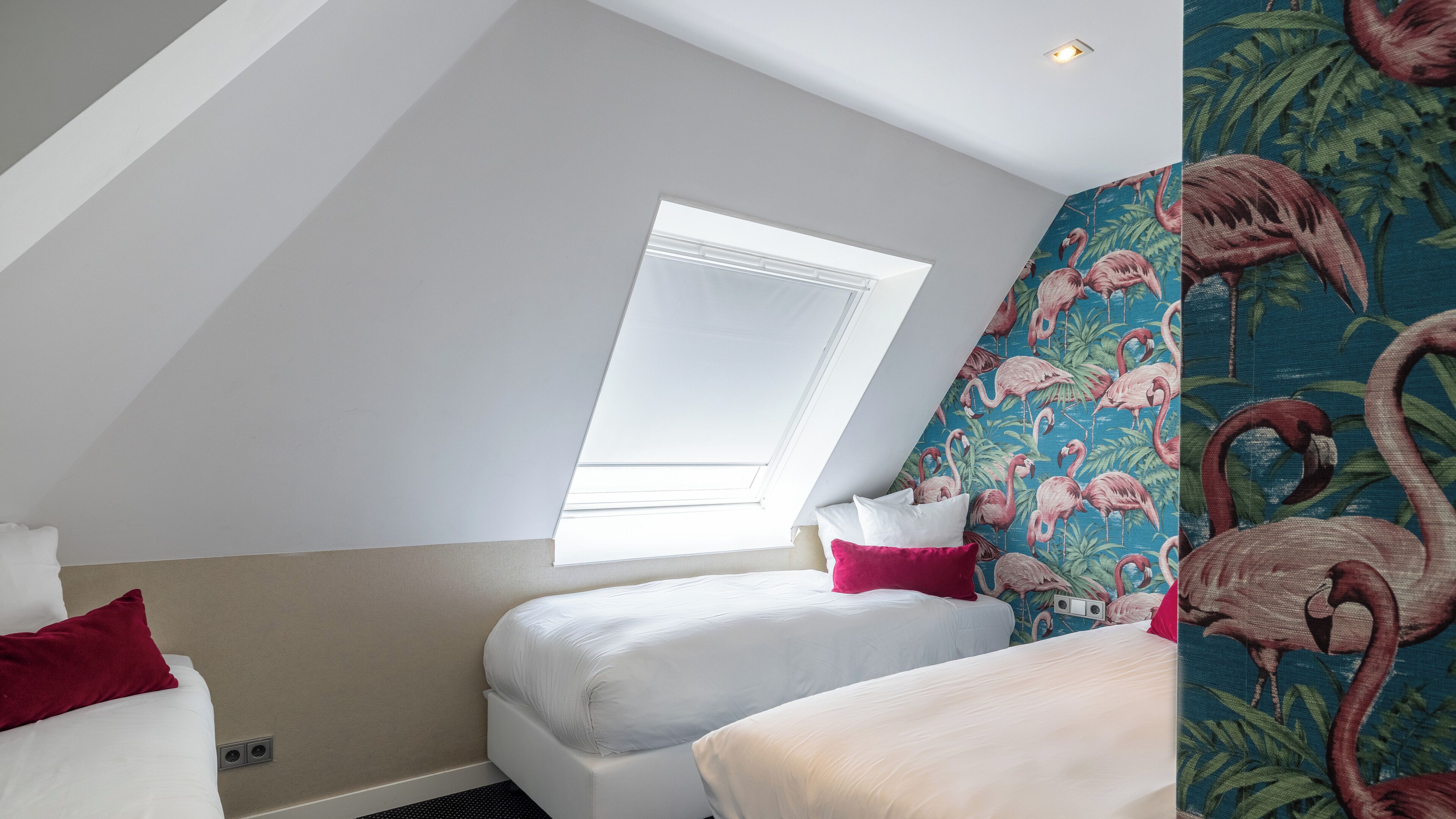 Quarto com claraboia, duas camas, almofada vermelha e parede com padrão de flamingos.