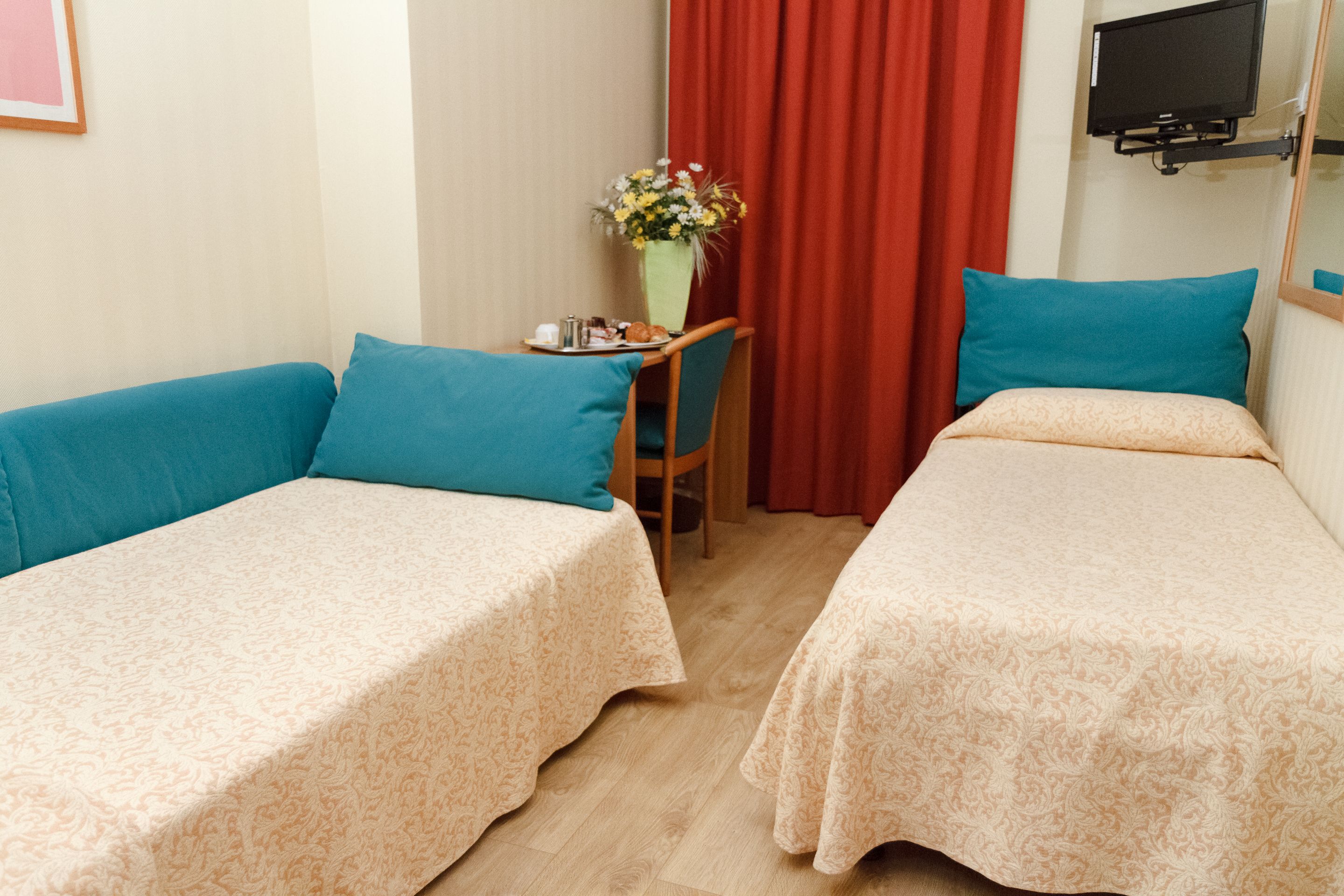 Quarto de hotel com duas camas, uma mesinha com um vaso de flores, uma cadeira, uma televisão e uma cortina vermelha.