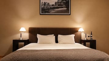 Quarto de hotel com uma cama grande, duas mesas de cabeceira com luminárias, um quadro na parede e uma garrafa de vinho sobre uma das mesas.