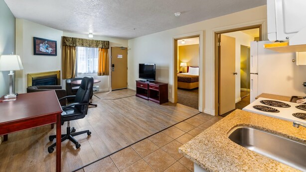 Quarto de hotel com piso de madeira, uma escrivaninha com luminária, televisão, sofá, mesa de jantar, e uma cozinha com fogão, além de uma cama no quarto adjacente.