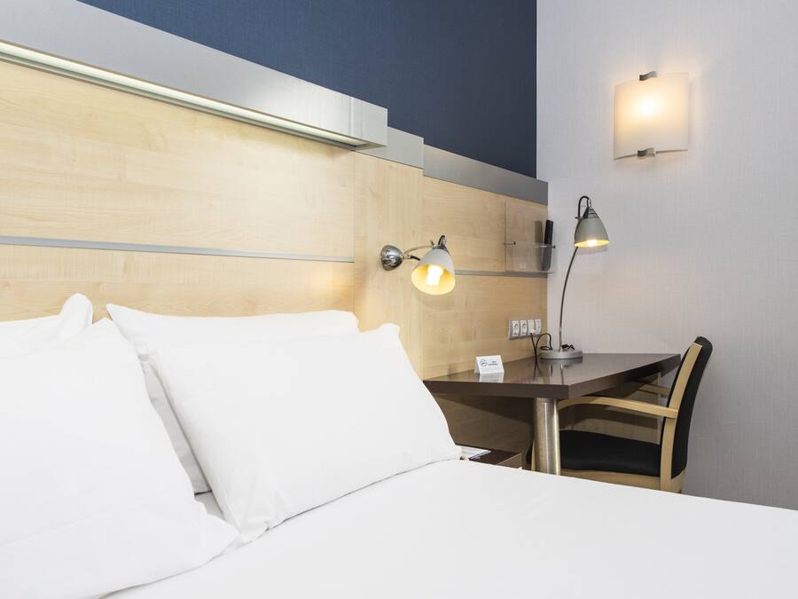 Quarto de hotel com uma cama, uma escrivaninha e duas luminárias fixadas na parede.