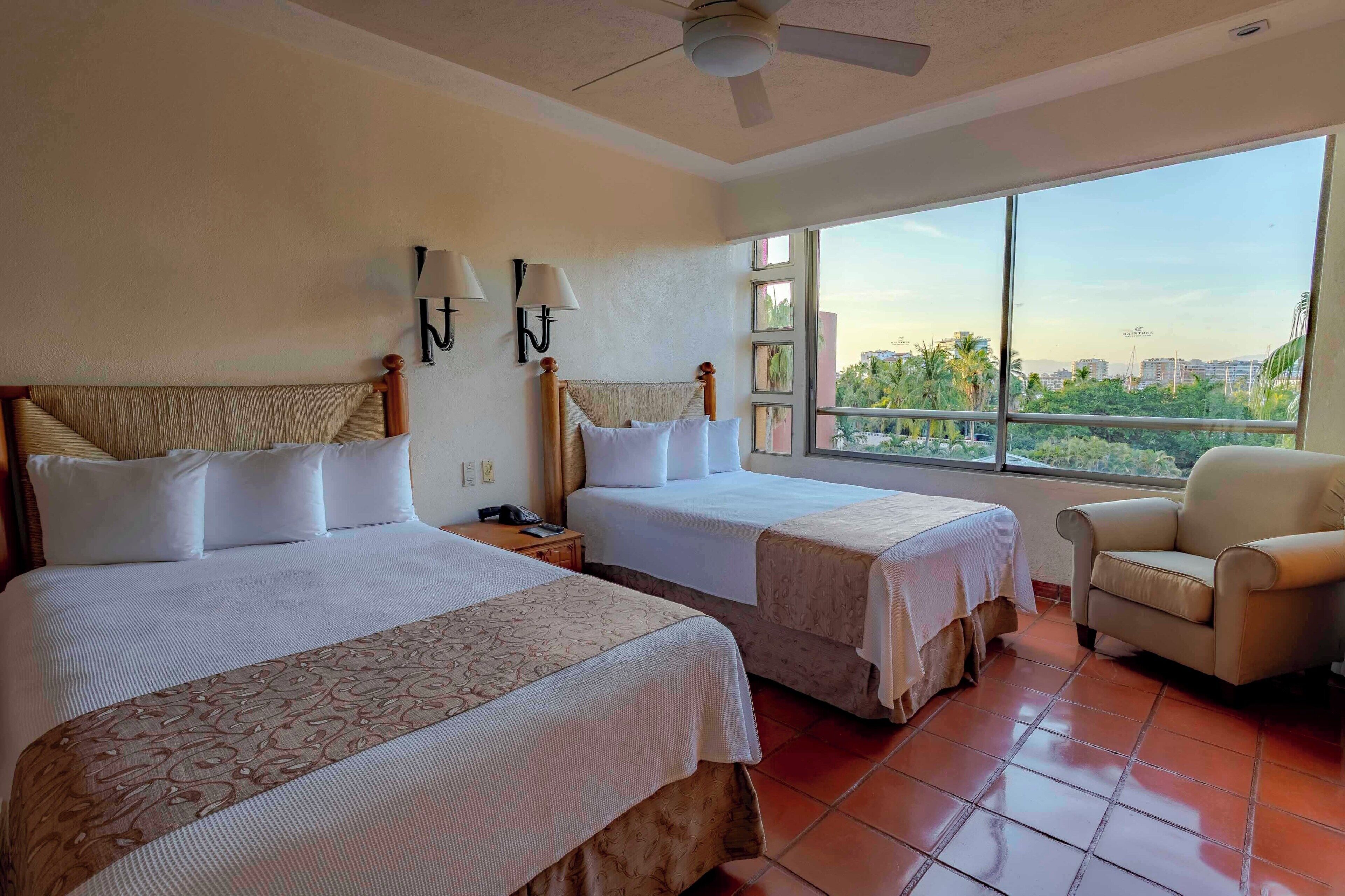 Quarto de hotel com duas camas, uma cadeira, ventoinha no teto e uma janela grande com vista para a cidade.