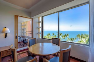 Quarto com uma grande janela que oferece vista para o oceano e palmeiras. Há uma mesa redonda com cadeiras, um abajur e uma porta que dá para outro quarto.