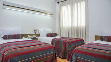 Quarto de hotel com duas camas, uma mesa de cabeceira com abajur e uma janela grande com cortinas.