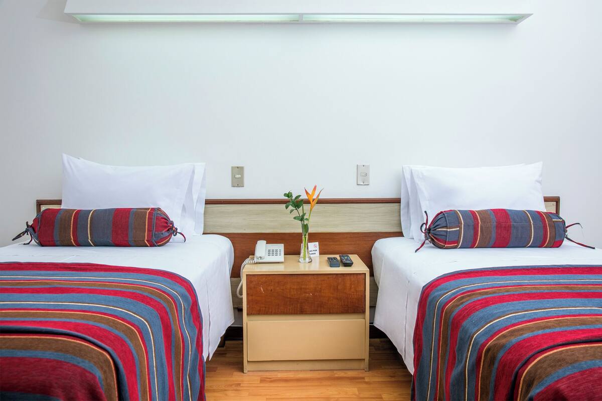 Quarto de hotel com duas camas, uma mesa de cabeceira com telefone, um vaso com flores e uma mesinha de cabeceira com controle remoto.