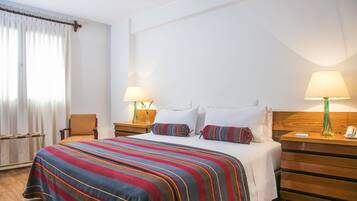 Quarto de hotel com uma cama grande, duas mesas de cabeceira, uma cadeira e uma janela com cortinas.