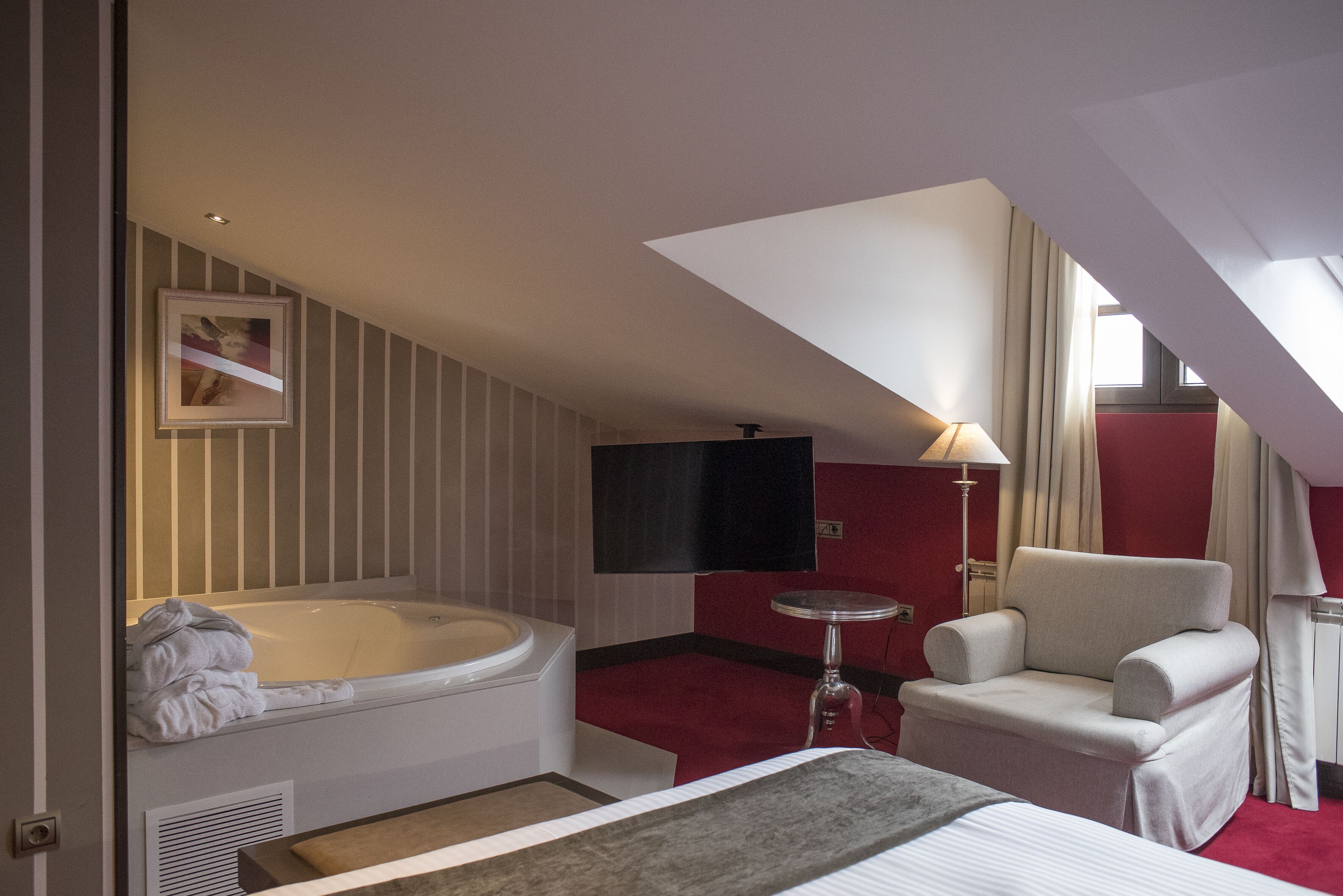 Quarto de hotel com uma banheira grande, uma poltrona confortável, uma televisão de tela plana e uma mesinha com abajur.