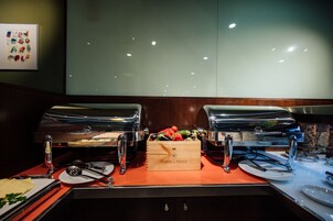 Disposição de buffet com duas estações de comida cobertas, uma caixa de madeira com a inscrição “QUADRO AL TASSO” e vários utensílios e pratos em um balcão.