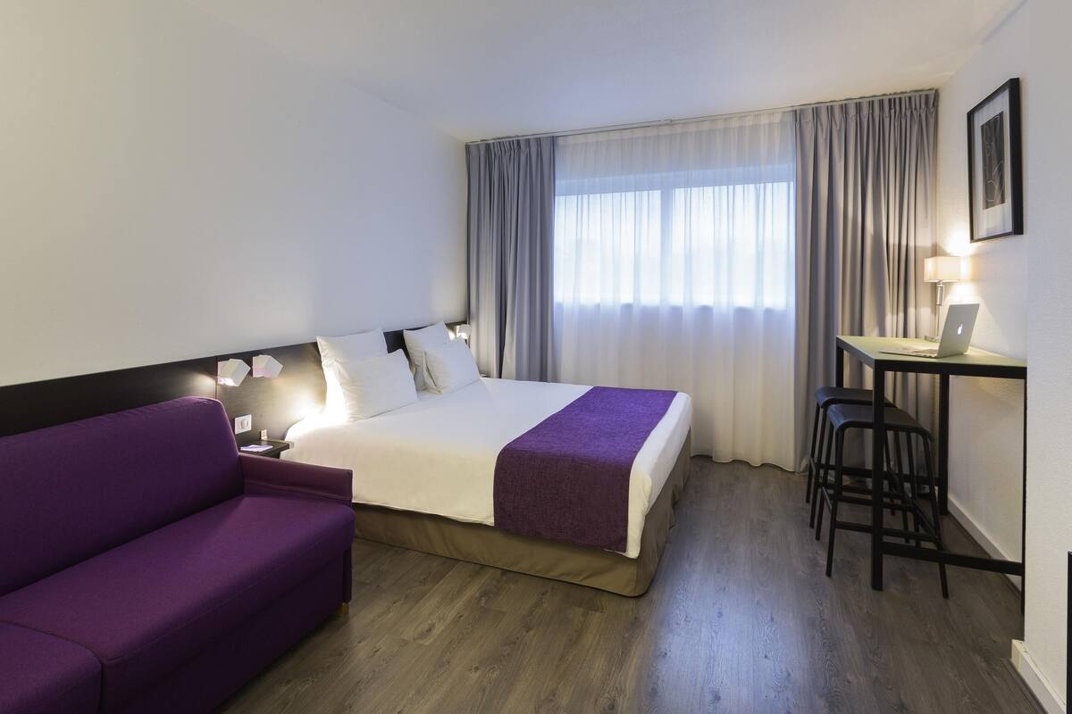 Quarto de hotel com uma cama, um sofá roxo, uma escrivaninha com um laptop e uma janela com cortinas.